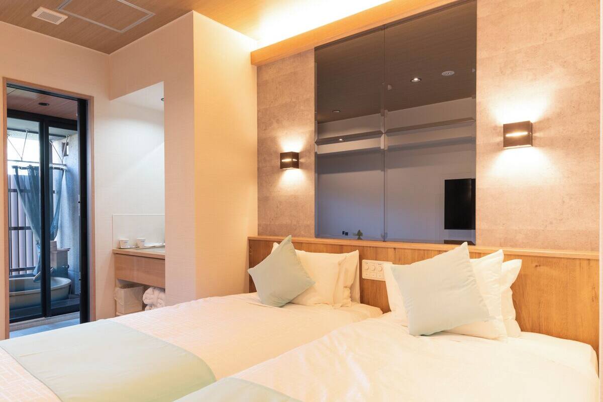 Quarto de hotel com duas camas, cabeceira de madeira, um espelho grande e porta de vidro deslizante para uma varanda.
