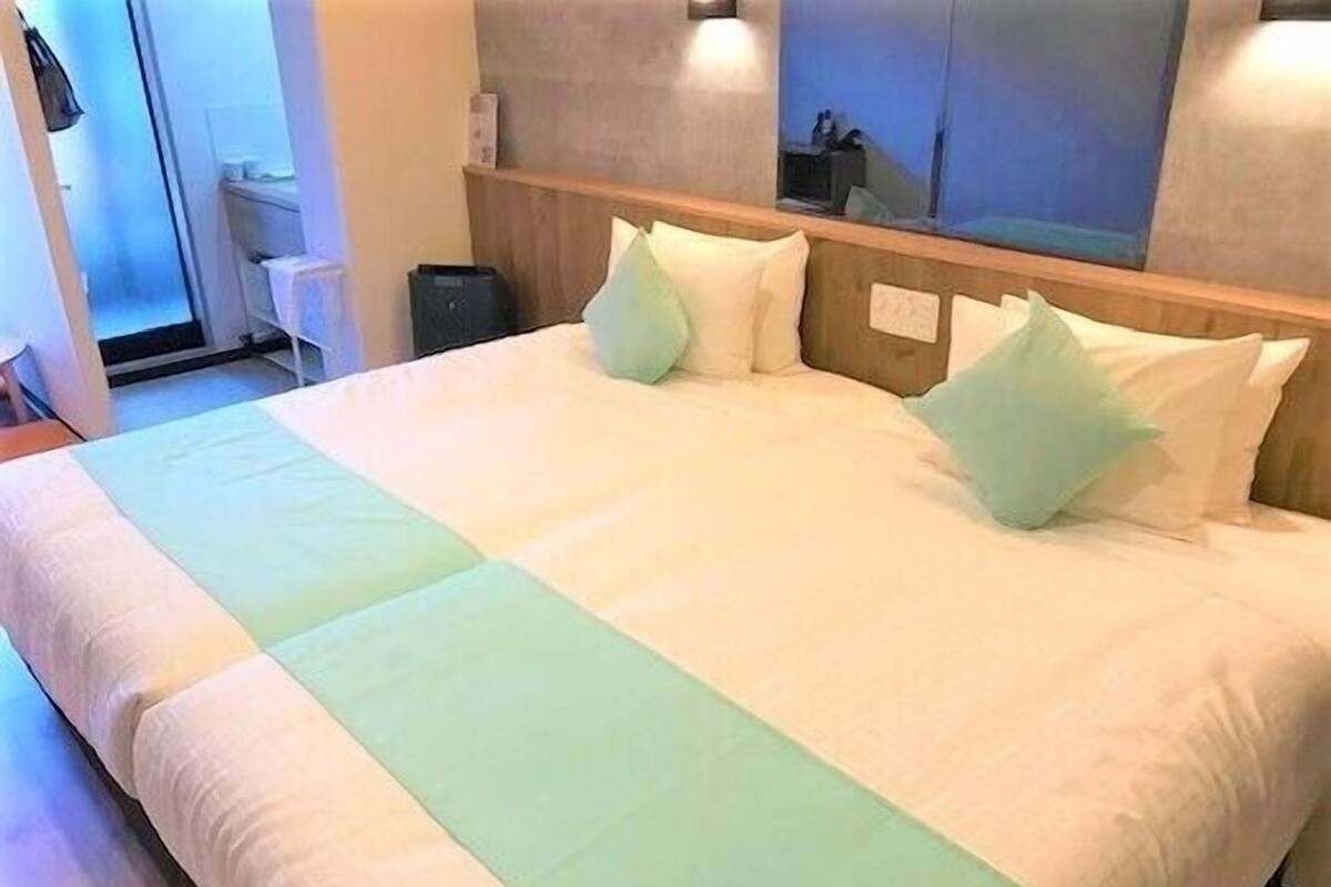 Uma cama bem arrumada com colcha branca e azul clara, cabeceira de madeira e uma pequena mesa de cabeceira com luminária.