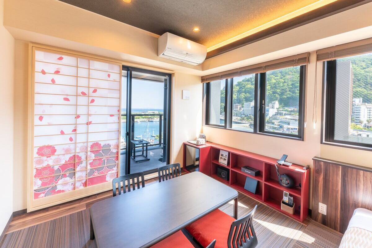 Quarto de hotel com uma mesa grande, cadeiras, um sofá vermelho, um painel deslizante shoji e vista da cidade através de amplas janelas.