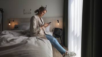 Uma pessoa sentada na cama de um quarto de hotel, usando um smartphone. O quarto tem um design moderno com cabeceira de madeira, duas luminárias de cabeceira e vista para a cidade pela janela.