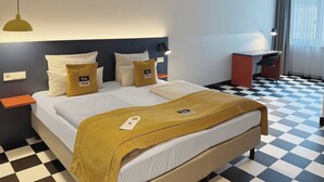 Quarto de hotel com uma cama, uma escrivaninha e uma cadeira. Há uma etiqueta de check-in na cama.