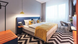 Quarto de hotel com cama, mesa de cabeceira, luminária, cadeira, escrivaninha e uma janela grande com cortinas.