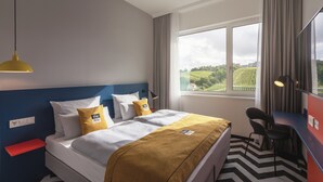 Quarto de hotel com cama, escrivaninha, cadeira, janela com vista para o verde e luminária pendente.