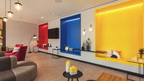 Um interior moderno com uma parede destacada vermelha, uma parede destacada azul e uma parede destacada amarela. Há uma TV de tela plana montada na parede vermelha, uma parede azul com uma prateleira, e uma parede amarela com uma prateleira. Existe uma cadeira vermelha, um sofá cinza e uma pequena mesa redonda com um vaso amarelo.