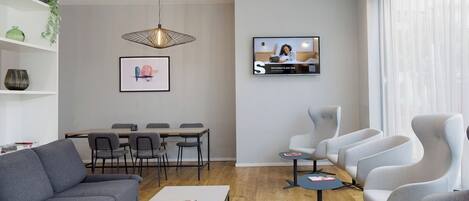 Quarto de hotel moderno com sofá, mesa de jantar com cadeiras, TV fixada na parede e uma janela grande com cortinas.
