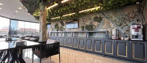 Uma área de bar moderna com parede de pedra, folhagem verde e uma máquina de Nescafé.