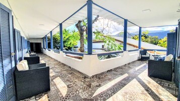 Uma área de estar coberta ao ar livre, com piso estampado, pilares azuis e vista para o verde e montanhas.