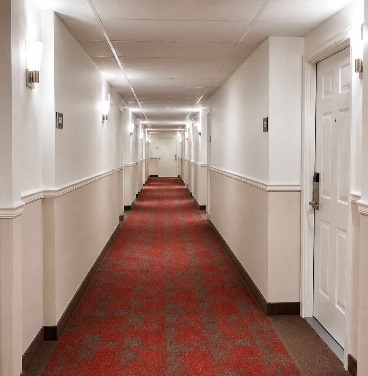 Um corredor de hotel com carpete vermelho, paredes brancas e portas em ambos os lados.
