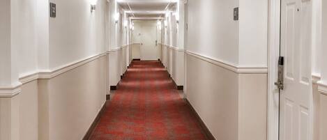 Um corredor de hotel com carpete vermelho, paredes brancas e portas em ambos os lados.