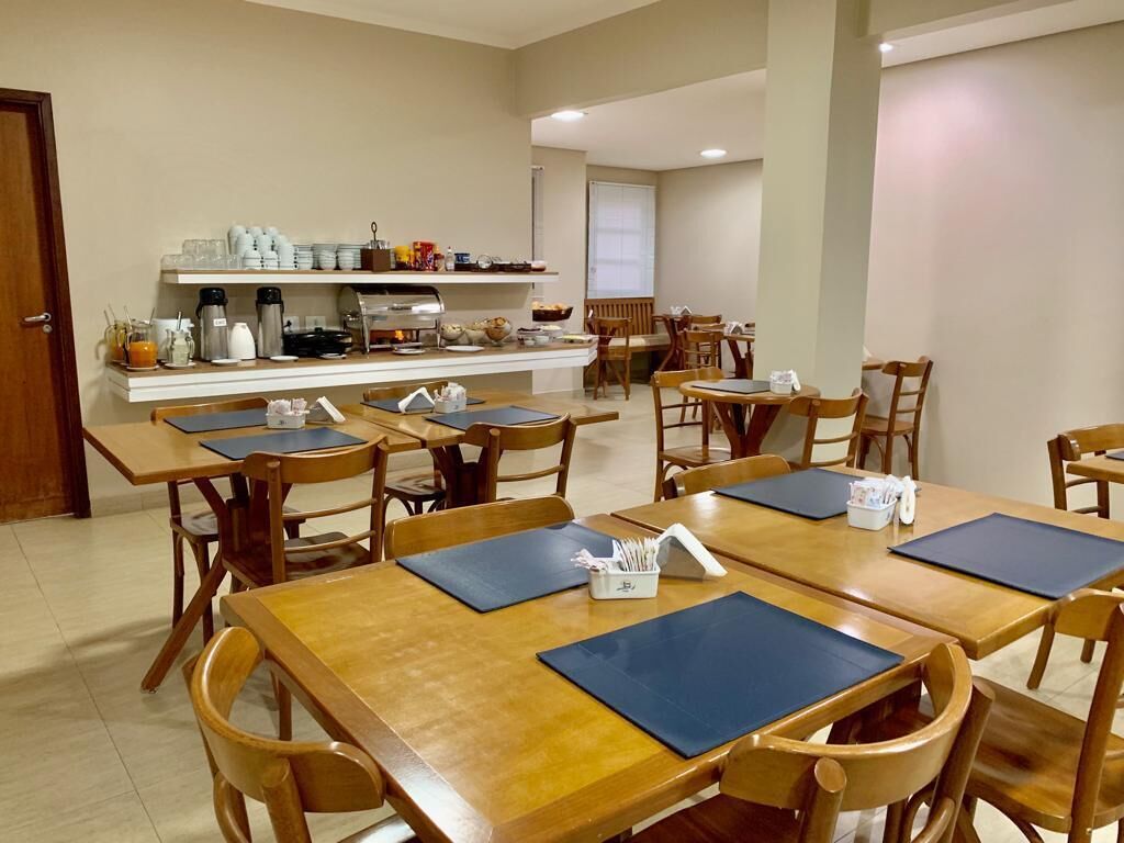 Uma área de refeições com mesas e cadeiras de madeira, um buffet com diversos alimentos e uma cozinha ao fundo.