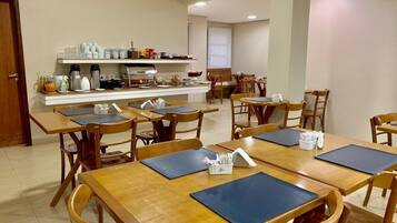 Uma área de refeições com mesas e cadeiras de madeira, um buffet com diversos alimentos e uma cozinha ao fundo.