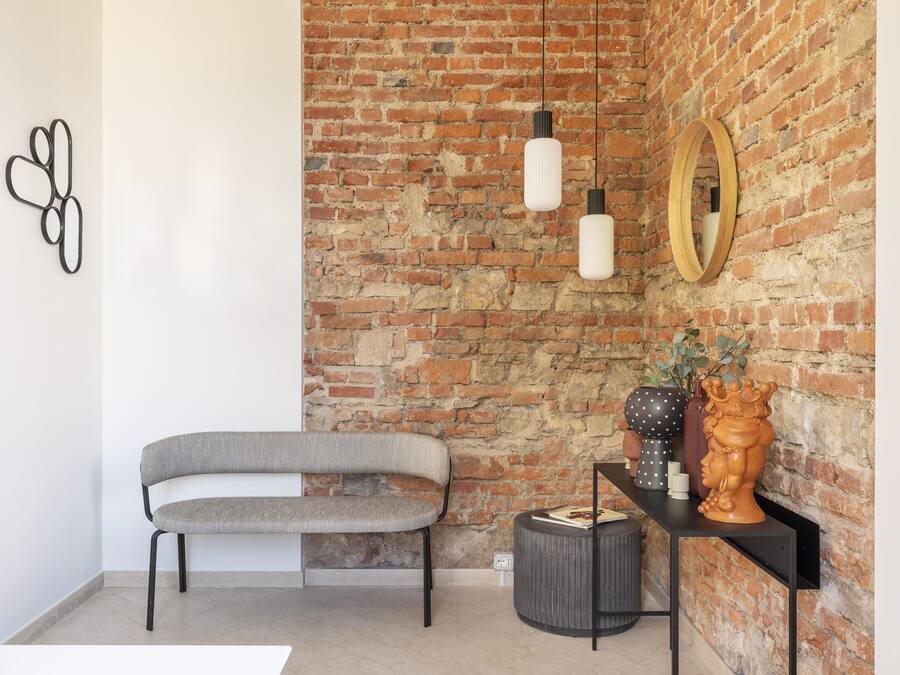 Quarto moderno com parede de tijolos, um banco cinza, mesa redonda com objetos decorativos e luminárias pendentes.