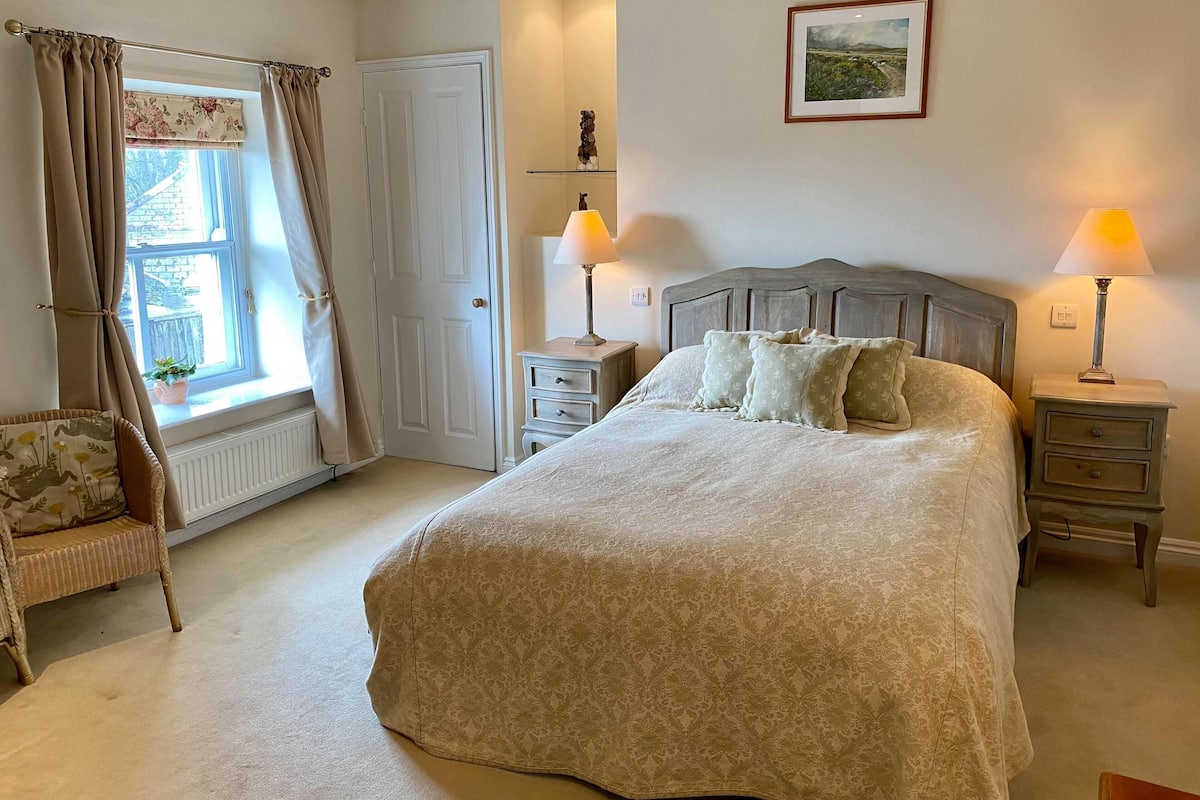 Um quarto com cama, duas mesas de cabeceira, uma cadeira, janela com cortinas, porta, prateleira na parede e um quadro emoldurado.
