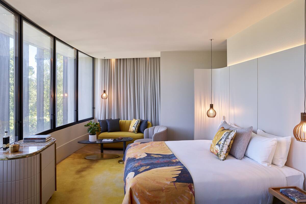 Quarto de hotel moderno com uma cama grande, um sofá, uma pequena mesa e uma escrivaninha. Janelas amplas oferecem luz natural e vista para o exterior.