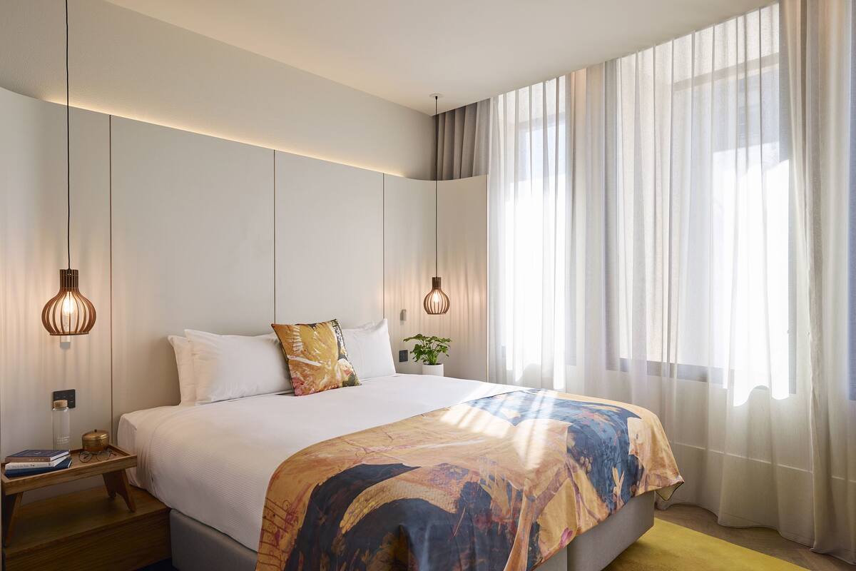 Quarto de hotel moderno com uma cama grande, duas luminárias pendentes, uma mesa de cabeceira com abajur e cortinas translúcidas.