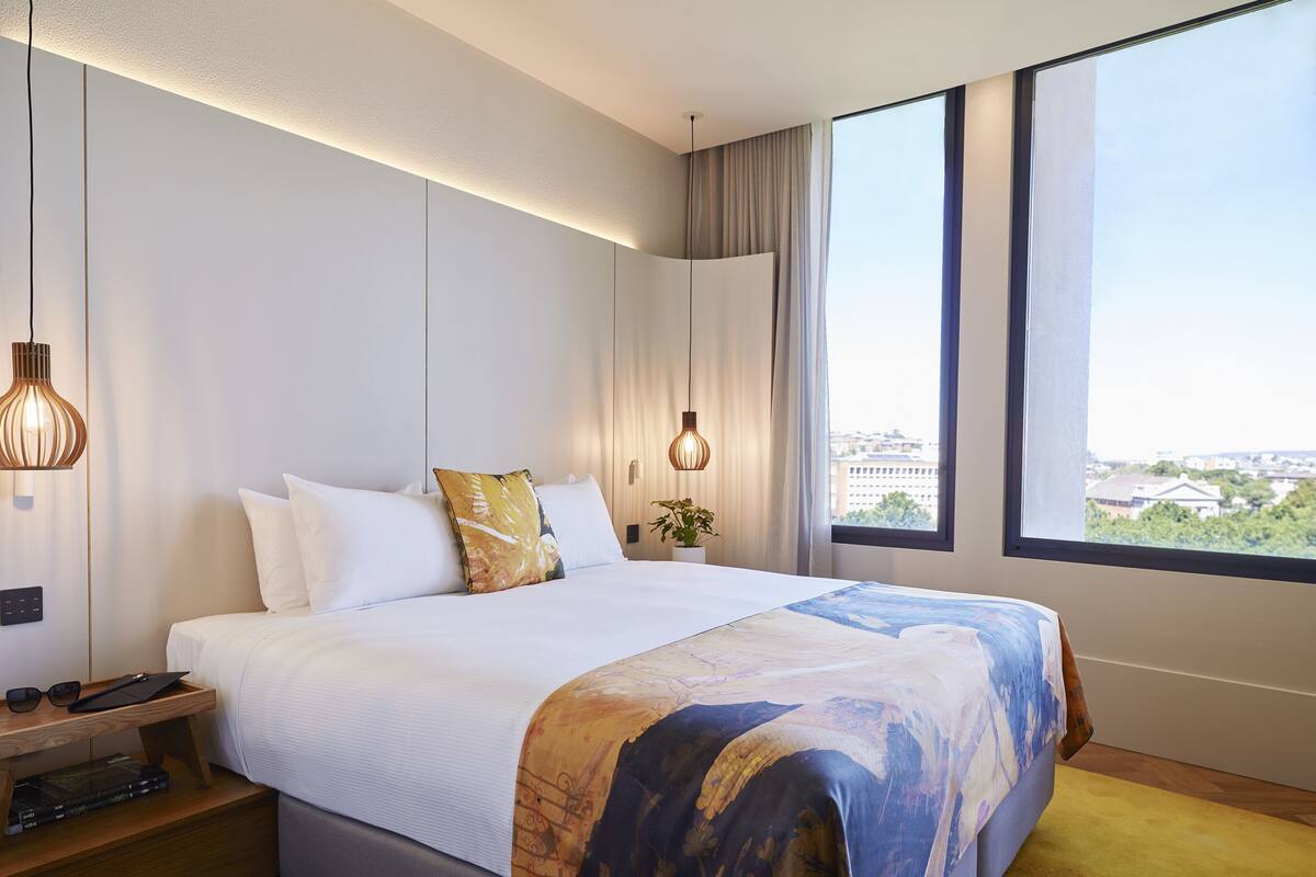 Quarto de hotel moderno com uma cama grande, mesas de cabeceira, vista para a cidade e uma janela ampla.