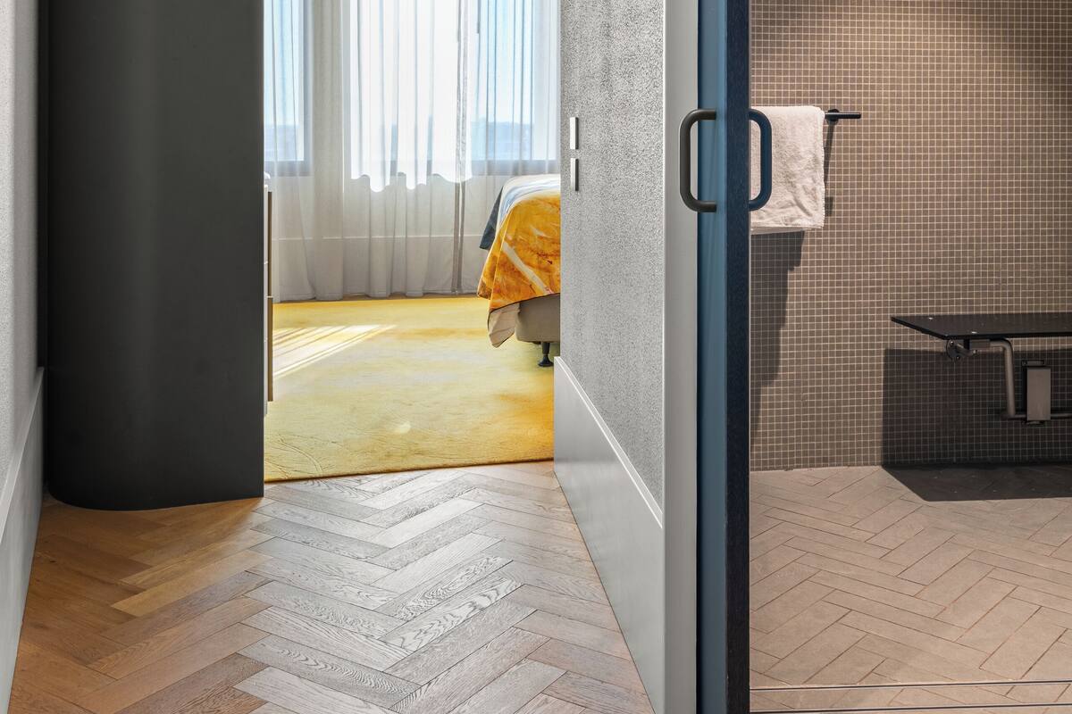 Quarto de hotel moderno com piso de madeira em padrão herringbone, um pilar escuro grande, uma porta de vidro deslizante que dá para um banheiro com parede de chuveiro revestida em azulejos, e uma cama com cabeceira amarela.