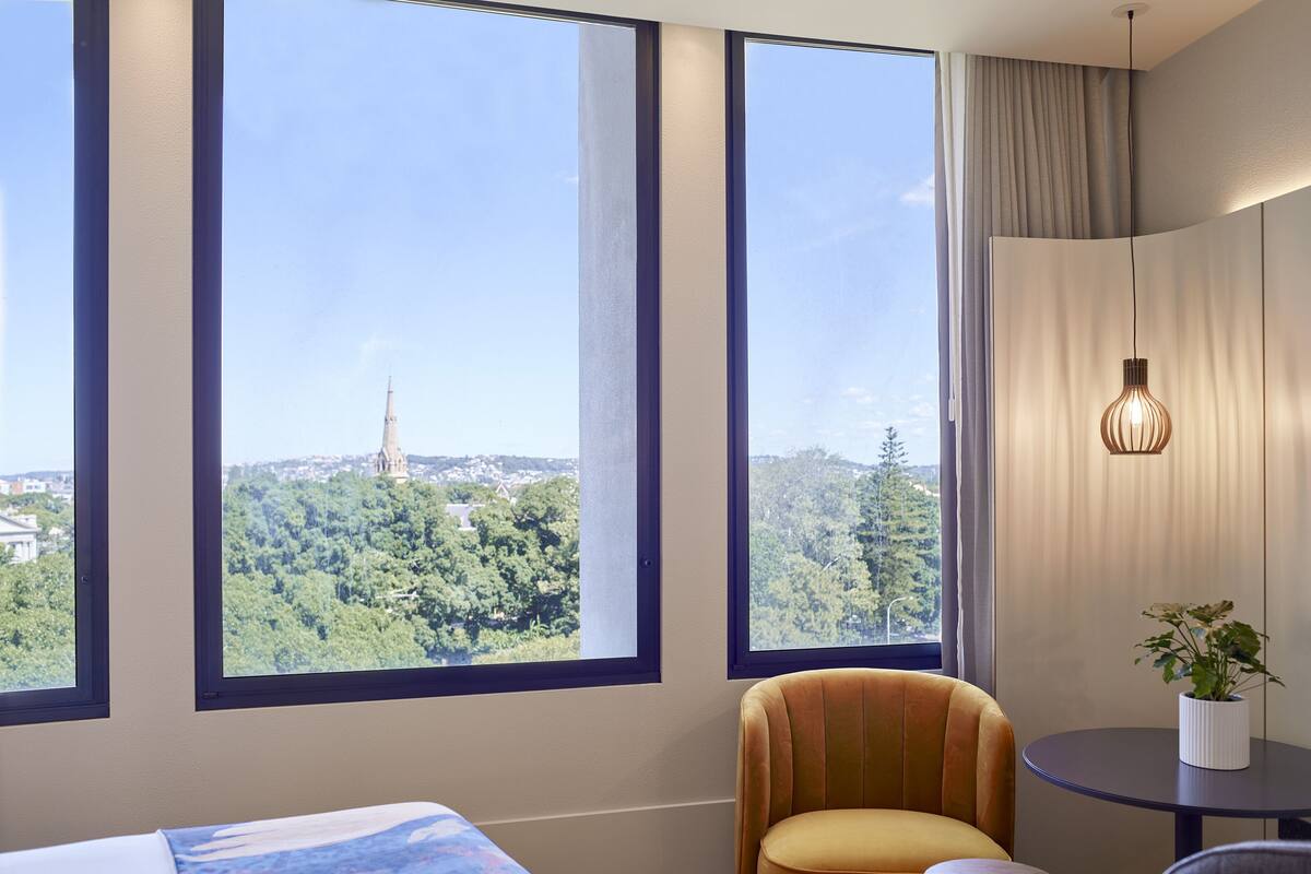 Quarto de hotel com grandes janelas, uma cama, uma cadeira, uma pequena mesa e um lustre.