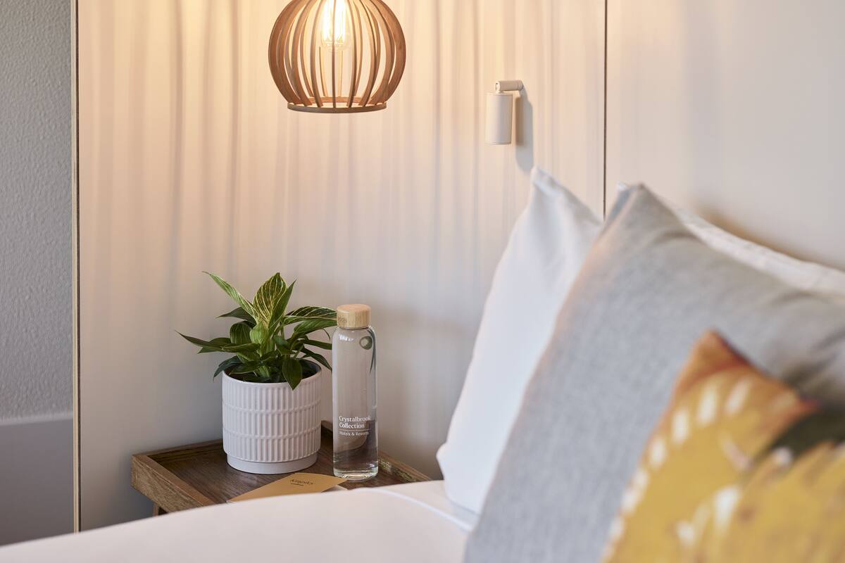 Quarto de hotel bem arrumado, com uma mesinha de cabeceira que exibe um vaso de planta, uma garrafa de água e um travesseiro branco.