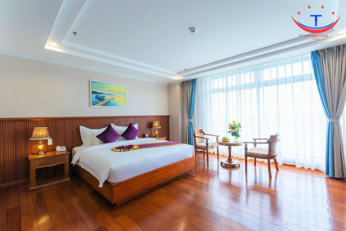 Quarto de hotel com uma cama grande, cabeceira de madeira, mesas de cabeceira com luminárias, uma pequena mesa com cadeiras e uma janela ampla com cortinas.