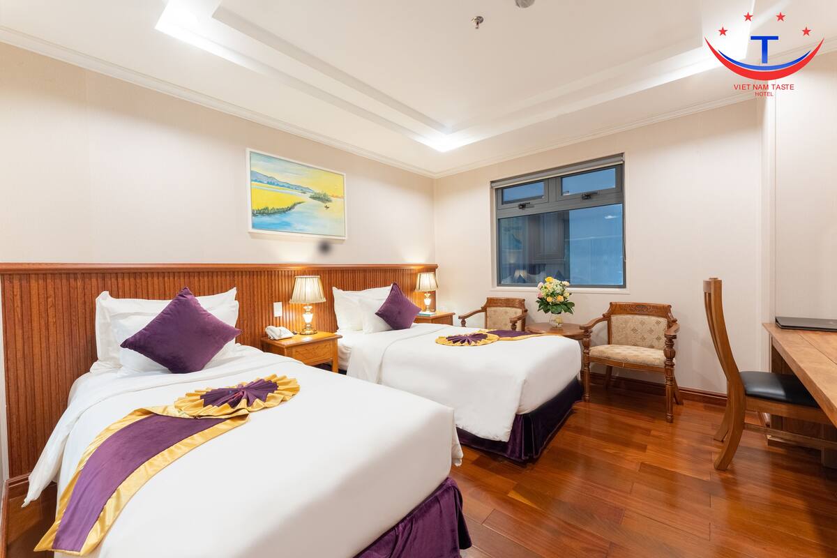 Quarto de hotel com duas camas, cabeceiras de madeira, uma escrivaninha, uma cadeira e uma janela com vista.