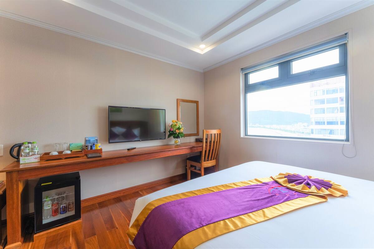 Quarto de hotel com cama, televisão, mini-geladeira, escrivaninha de madeira e janela com vista para edifícios.