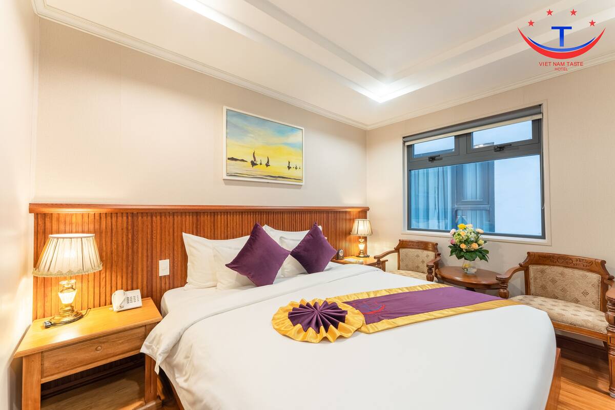Quarto de hotel com cabeceira de madeira, cama com roupa de cama branca, almofadas roxas, mesa de cabeceira com abajur, telefone, vaso de flores e janela com vista.