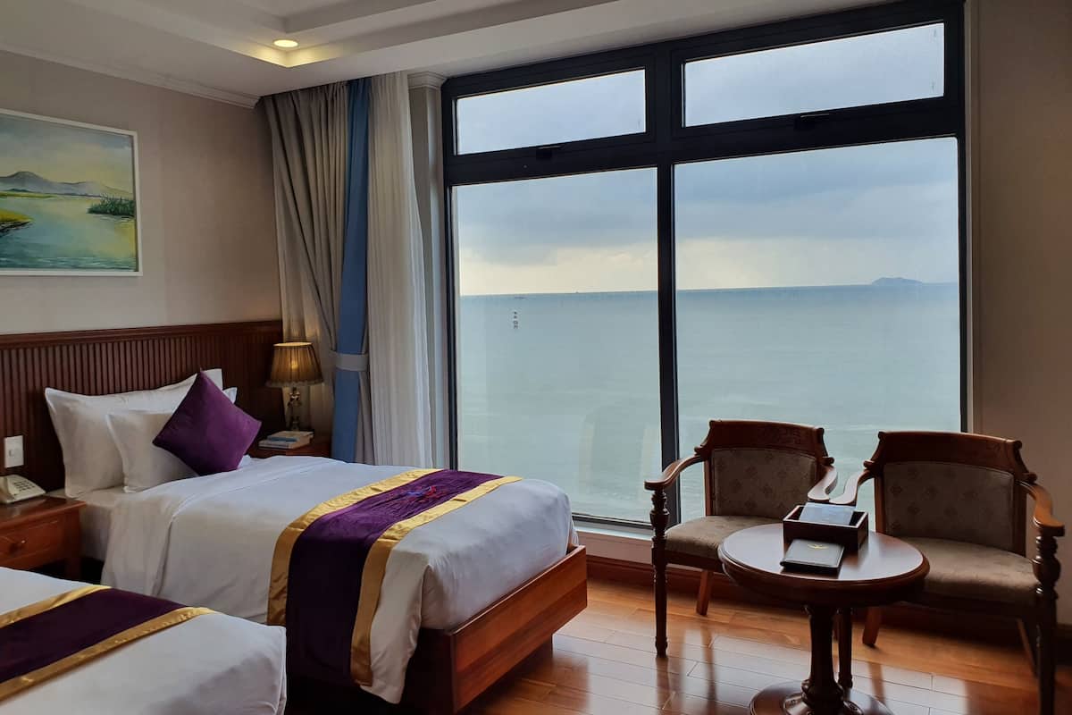 Quarto de hotel com uma janela grande que oferece vista para o mar, uma cama com colcha roxa e dourada, uma mesa de cabeceira com abajur e uma pequena mesa redonda com cadeiras.