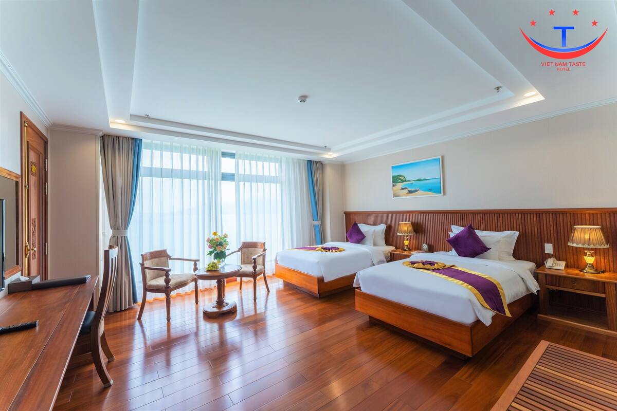 Quarto de hotel com duas camas, uma escrivaninha de madeira, uma cadeira, uma mesinha com flores e um quadro de uma praia na parede.