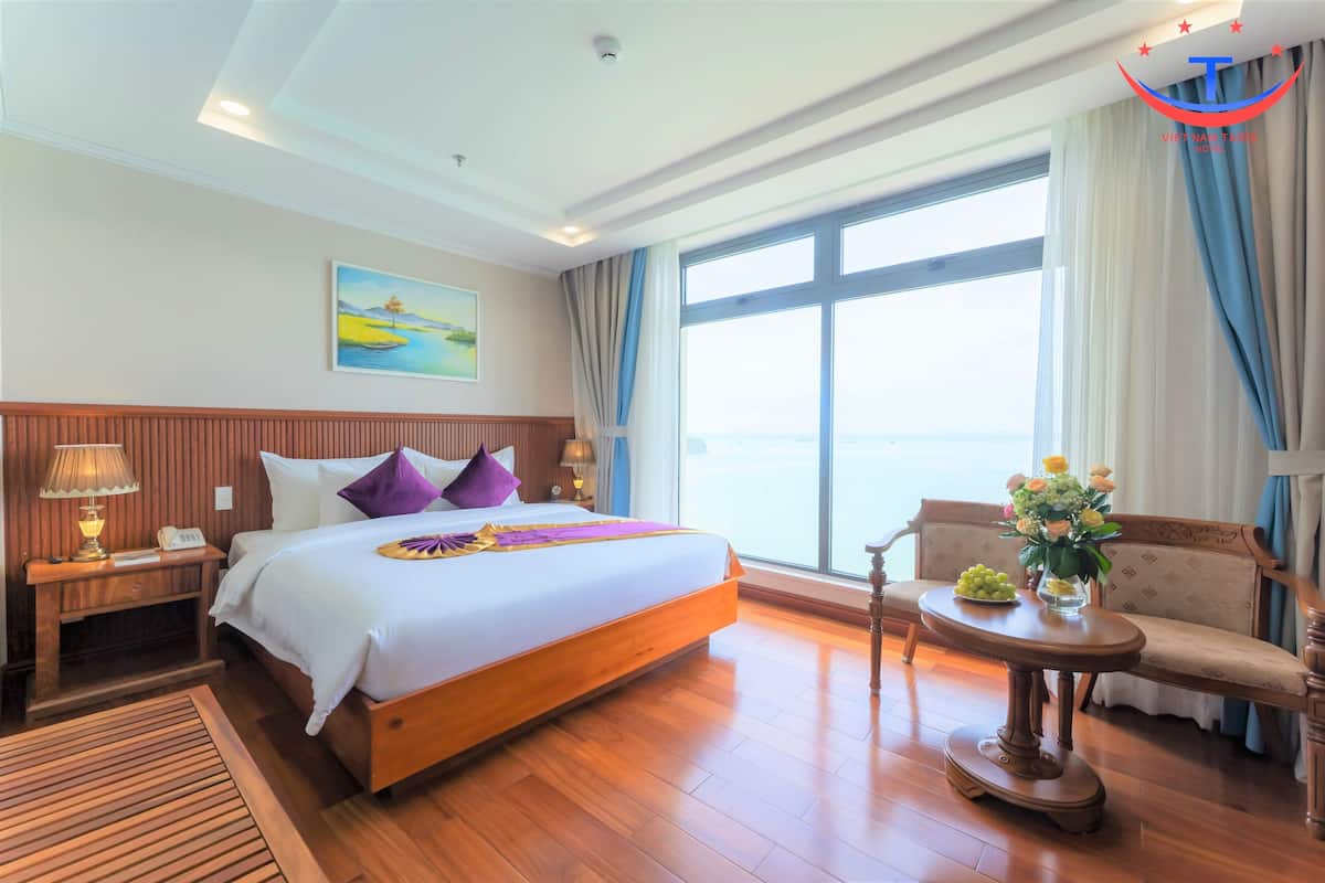 Quarto de hotel com cama grande, cabeceira de madeira, mesa de cabeceira com abajur, sofá, mesa redonda com um vaso de flores e uma janela grande com cortinas.