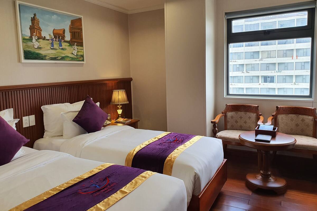 Quarto de hotel com duas camas, mesa de cabeceira, abajur, cadeira e janela com vista para edifícios.