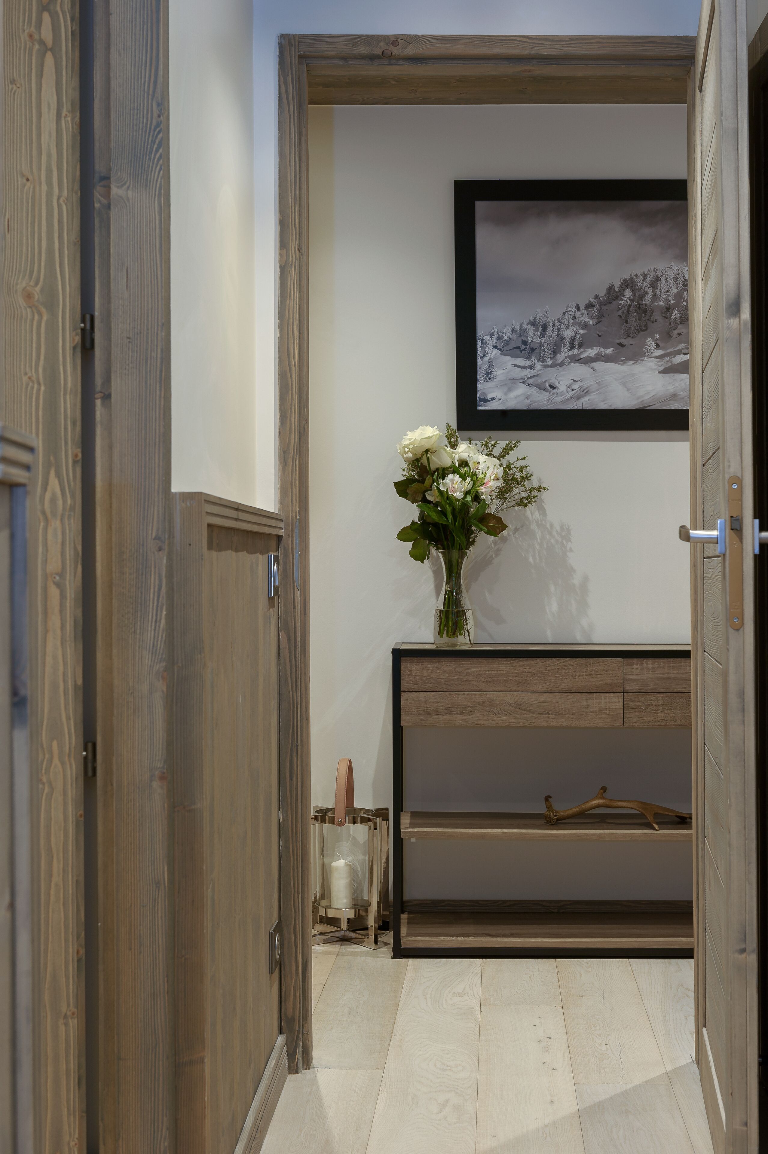 Um corredor com portas de madeira, um vaso de flores brancas, um quadro na parede e um aparador de madeira.