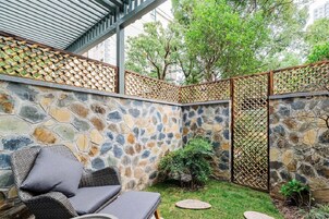 Uma área de estar ao ar livre com paredes de pedra, teto coberto, uma poltrona de vime e um pequeno jardim com cerca de bambu.
