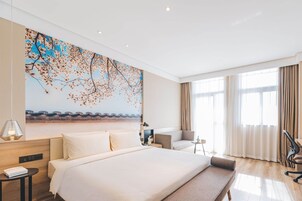 Quarto de hotel moderno com uma cama grande, uma escrivaninha, uma cadeira e uma janela ampla com cortinas.