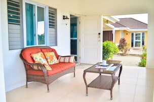 Um sofá de vime com almofadas coloridas, uma mesa com tampo de vidro e vista para uma casa através de uma porta aberta.