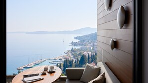 Quarto moderno com vista para uma vila costeira, uma mesa de madeira redonda com xícara e pires, e um sofá com almofadas.