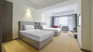 Quarto de hotel com duas camas, uma escrivaninha, uma cadeira e uma janela grande com cortinas.