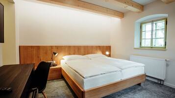 Quarto com cabeceira de madeira, cama com roupa de cama branca, escrivaninha com abajur e janela com vista para árvores.