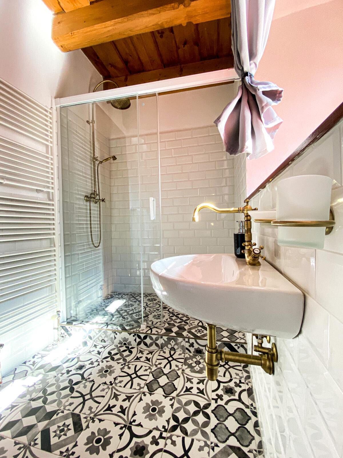 Casa de banho com chuveiro em cabine de vidro, lavatório branco com detalhes em latão e azulejos estampados.