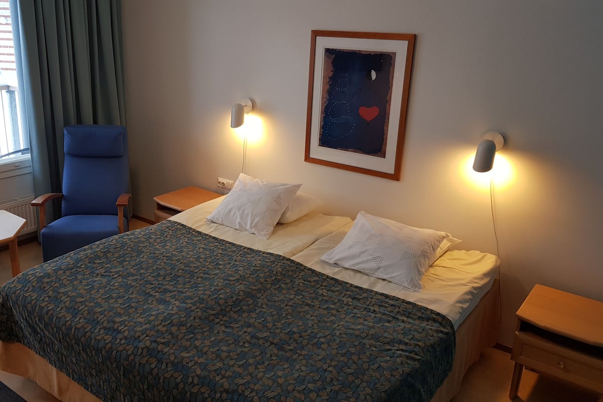 Quarto de hotel com cama, poltrona, mesa e janela com cortinas.