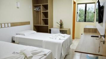 Quarto de hotel com duas camas, um guarda-roupa de madeira, uma mesa pequena com um vaso e uma janela com vista para o verde.
