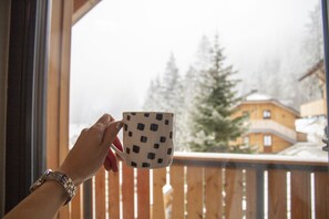 Uma mão segurando uma caneca com bolinhas, com um corrimão de madeira e uma paisagem nevada visível pela janela.