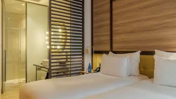 Quarto de hotel com cama grande, box de vidro, cabeceira de madeira e mesa de cabeceira com telefone e garrafa.
