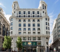Um edifício grande e branco, de vários andares, com arquitetura clássica, apresentando uma entrada central com toldo verde e diversas varandas.