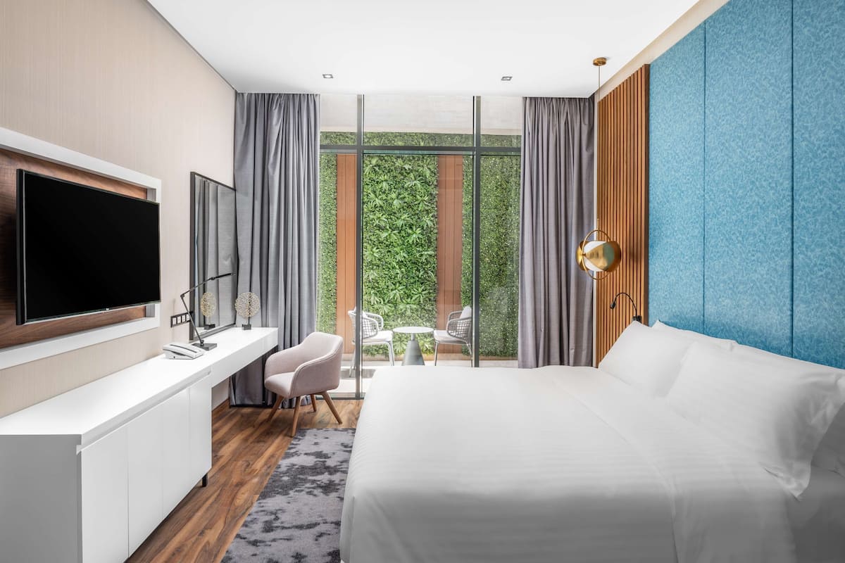 Quarto de hotel moderno com uma cama grande, uma escrivaninha com televisão de tela plana, uma pequena mesa com cadeiras e uma grande janela com cortinas.