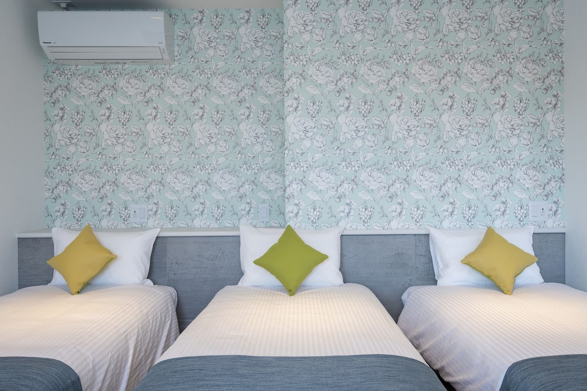 Quarto de hotel com três camas, cada uma com colcha branca e almofada. O quarto tem papel de parede floral e um ar condicionado na parede.