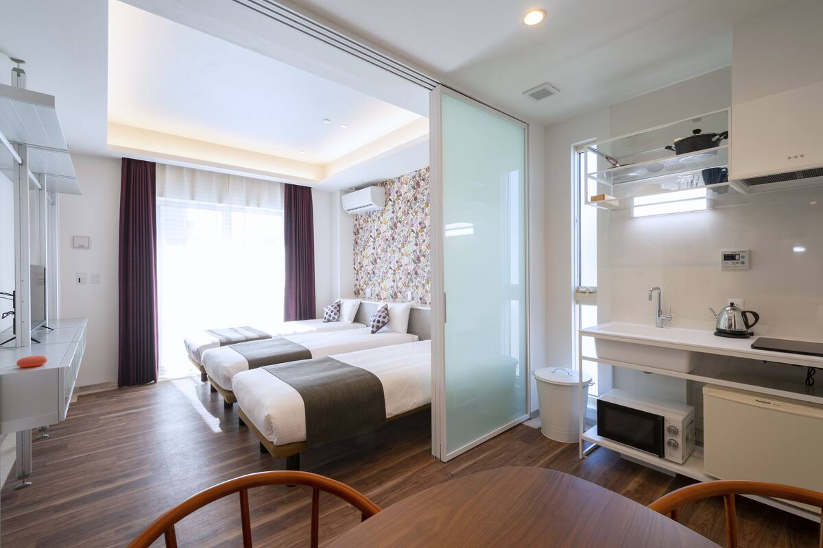 Quarto de hotel moderno com duas camas, uma escrivaninha, micro-ondas e porta de vidro deslizante para uma varanda.