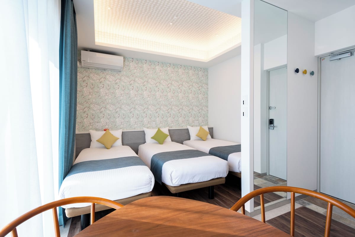 Quarto de hotel com três camas, uma mesa de jantar de madeira e uma porta de vidro que dá para um banheiro.