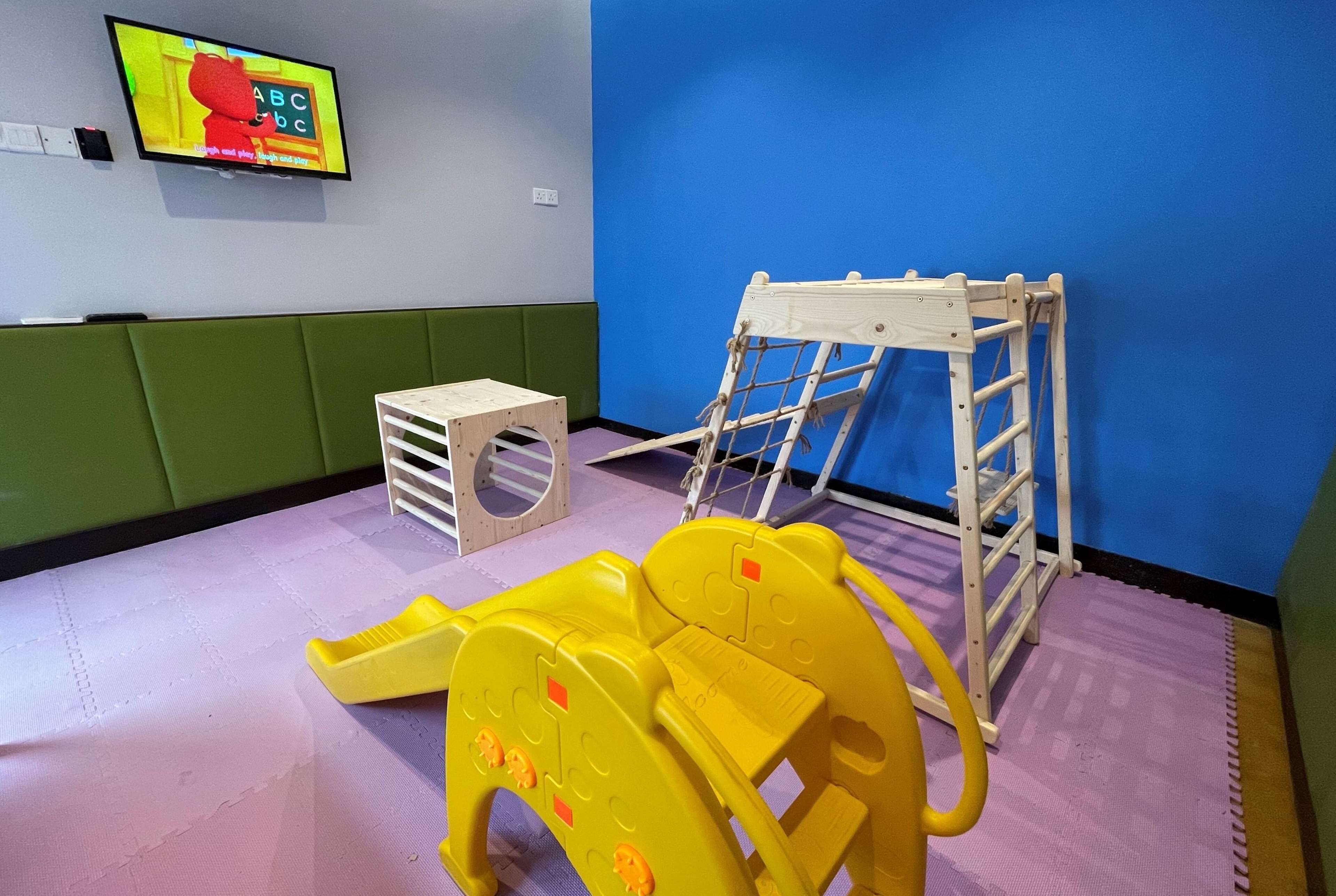 Uma sala de recreação com televisão, um banquinho pequeno, uma estrutura de brincadeira amarela e uma escada de escalada branca.