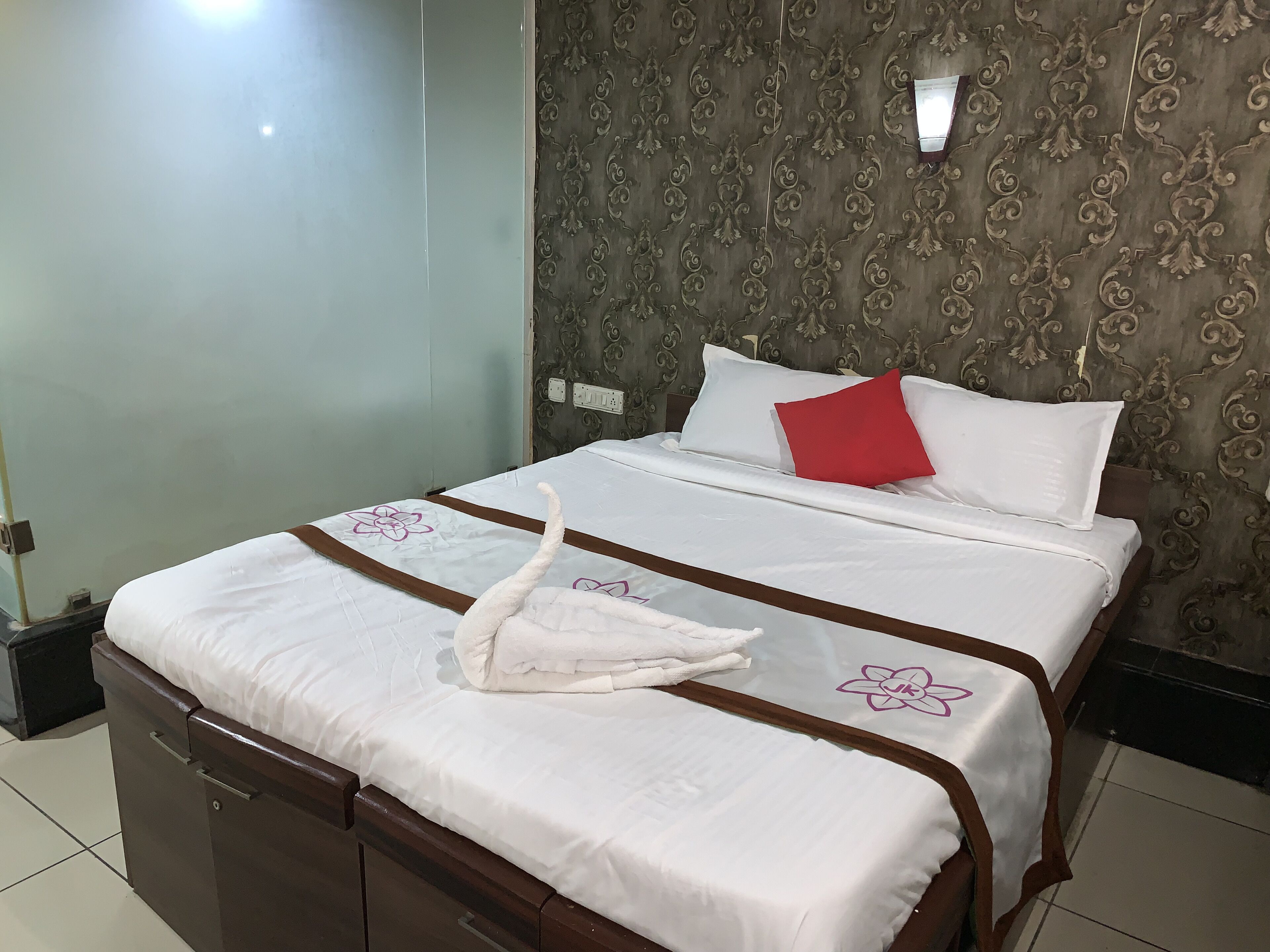 Uma cama bem arrumada com roupa de cama branca e um travesseiro vermelho, uma mesa de cabeceira de madeira com uma luminária, e uma parede com papel de parede decorado.